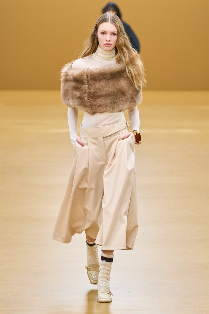 Runway model in cream turtleneck top with fur stole and wide-leg culotte trousers, styled with socks and loafers on minimalist runway backdrop