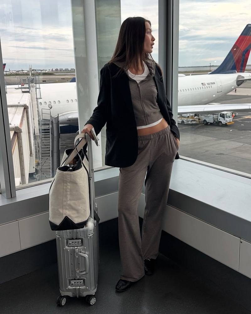 Traveler in relaxed lounge set with zip hoodie%2C blazer%2C and wide-leg pants%2C carrying tote and suitcase at airport window lounge