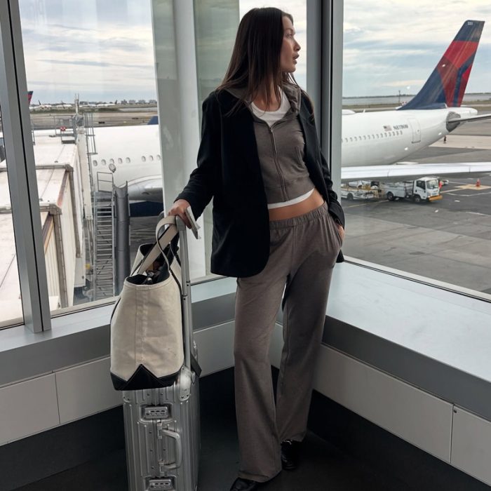Woman in airport lounge by a window, dressed in casual loungewear, holding a white tote and standing by a silver suitcase. Delta plane outside.