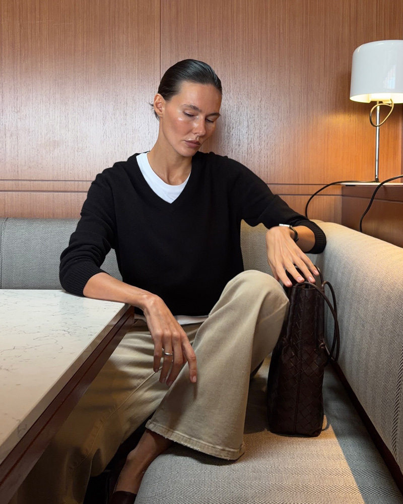 Woman in black knit sweater and cream trousers with woven leather tote bag%2C seated in a modern wood-paneled caf%C3%A9 setting