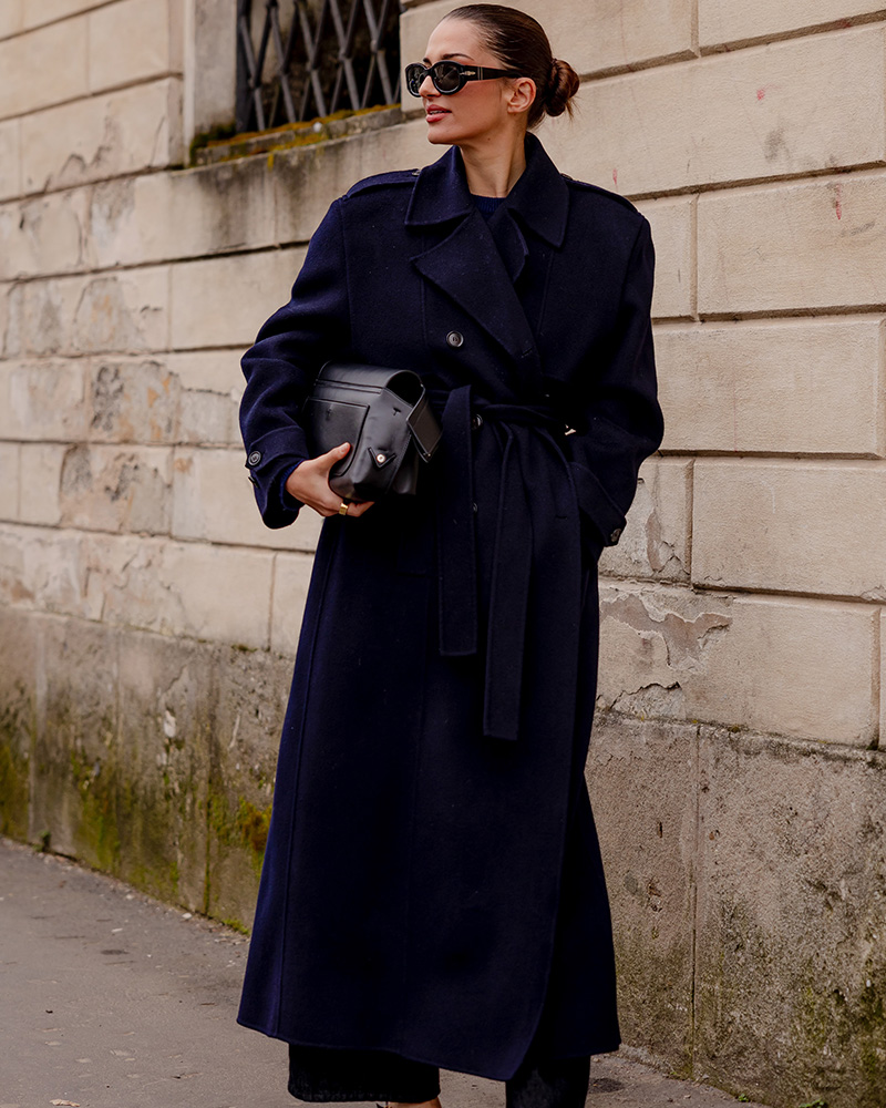 Street style featuring a navy belted maxi coat%2C black leather bag%2C sunglasses and pointed pumps against a stone wall%0A