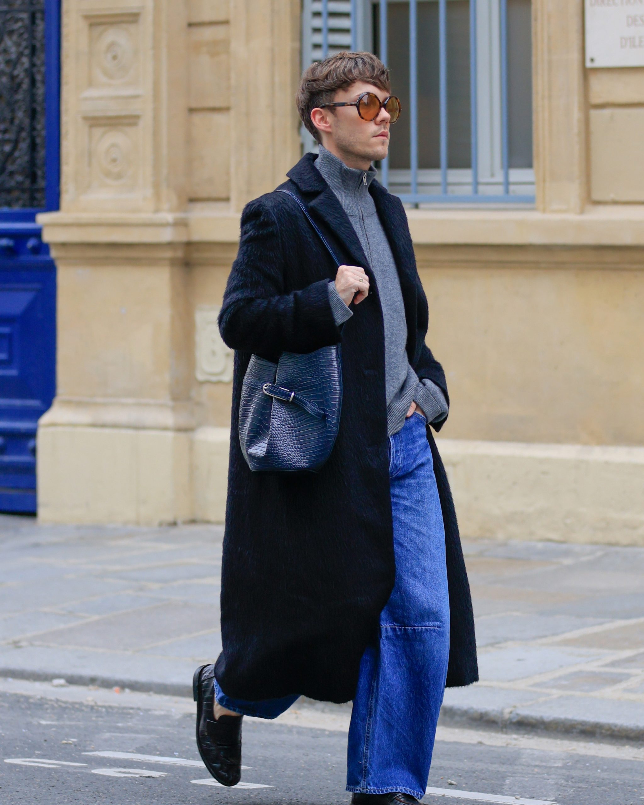 A stylish individual walks casually in a black oversized coat%2C gray turtleneck%2C wide-leg jeans%2C and black loafers%2C complemented by oversized sunglasses and a textured blue bag.