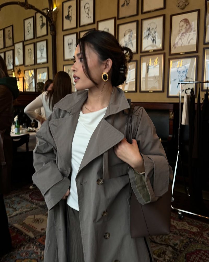 Taupe trench coat layered over white tee with gold hoop earrings and leather shoulder bag in a classic caf%C3%A9 setting