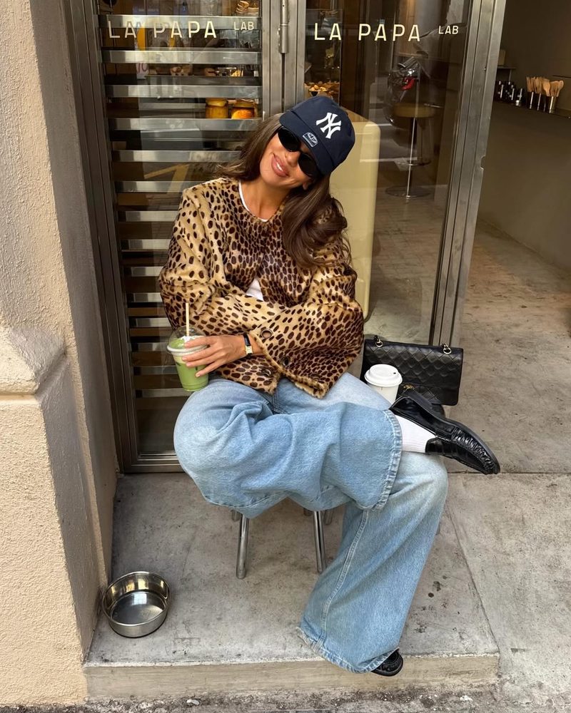 Stylish woman seated outside a caf%C3%A9%2C wearing a leopard print oversized cardigan%2C baggy light-wash jeans%2C and black loafers%2C complemented by a New York Yankees cap and sunglasses Stylish woman seated outside a caf%C3%A9%2C wearing a leopard print oversized cardigan%2C baggy light-wash jeans%2C and black loafers%2C complemented by a New York Yankees cap and sunglasses