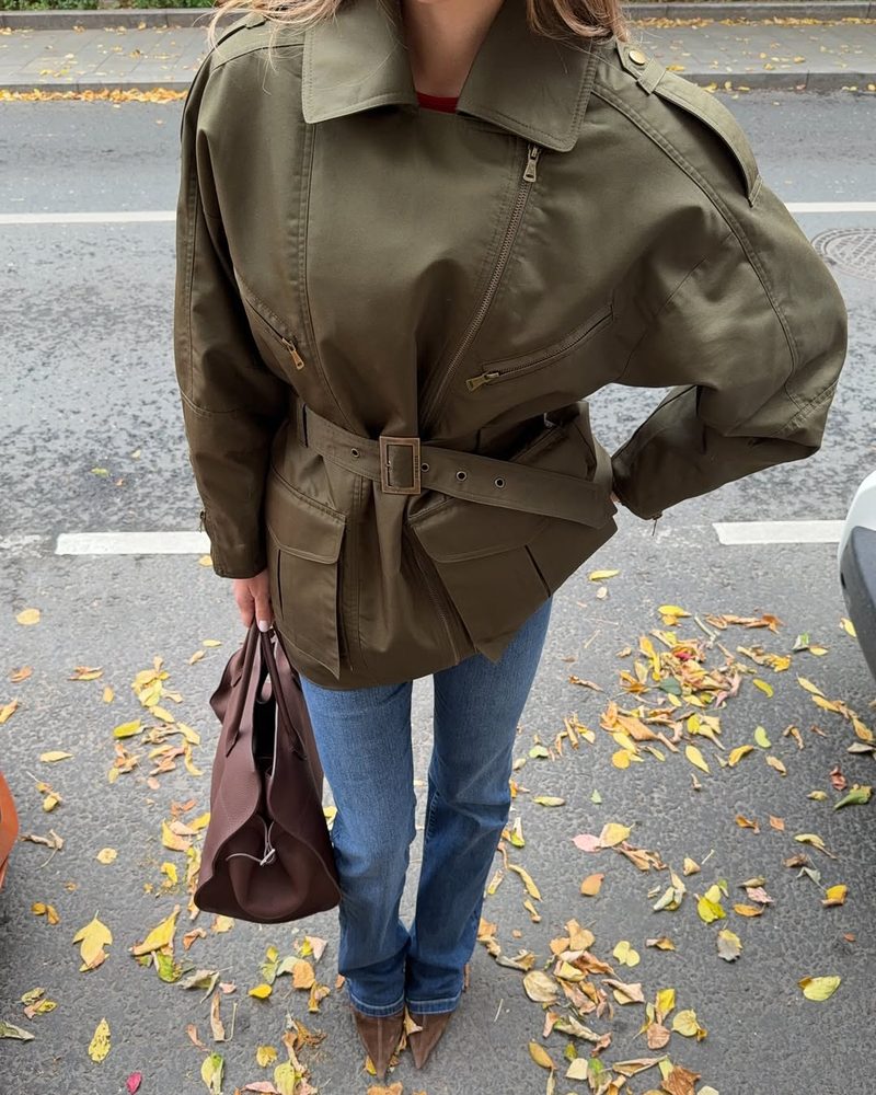 Stylish outfit featuring an olive green parka with a belted waist%2C paired with light blue flared jeans and a brown tote bag%2C set against a fall backdrop