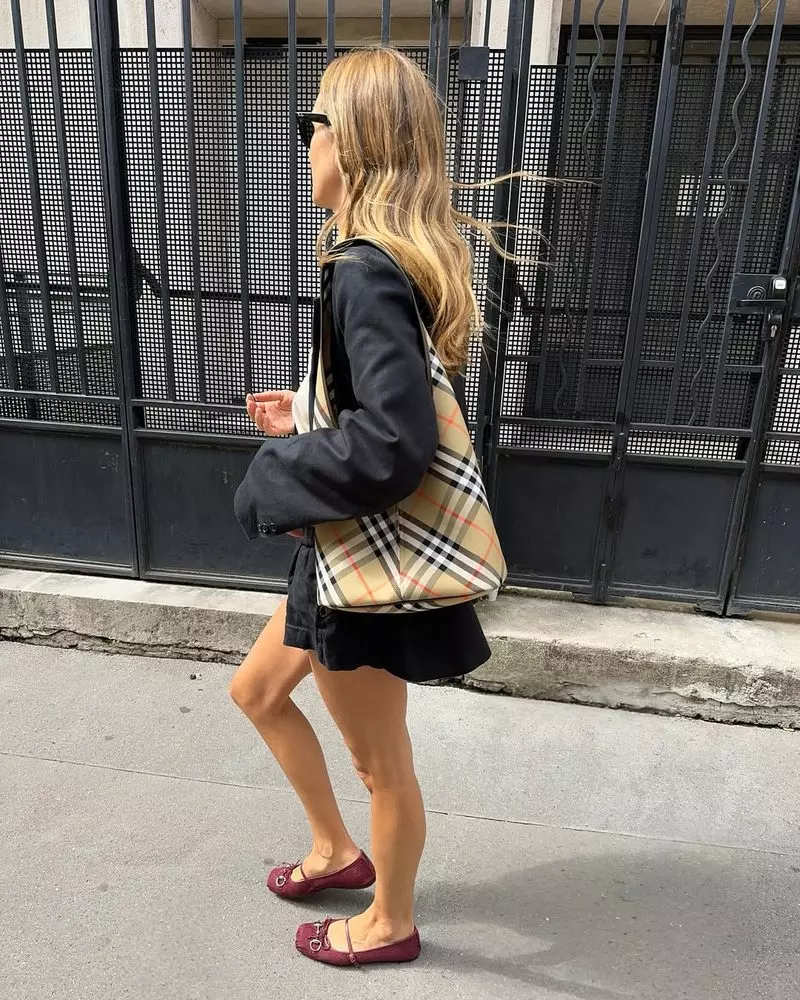 Woman walking on a city sidewalk wearing a black blazer%2C black mini dress%2C and burgundy flats with buckle detail%2C accessorized with a large Burberry check tote bag and black sunglasses.