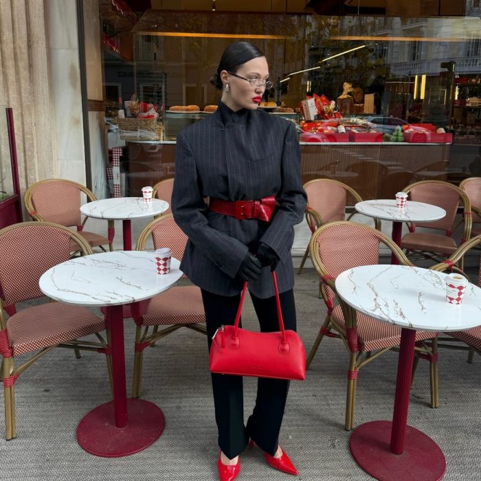 Chic woman styled in a pinstripe blazer with a bold red belt, paired with tailored black pants, red pointed-toe heels, and a matching handbag, set against a vibrant café backdrop.