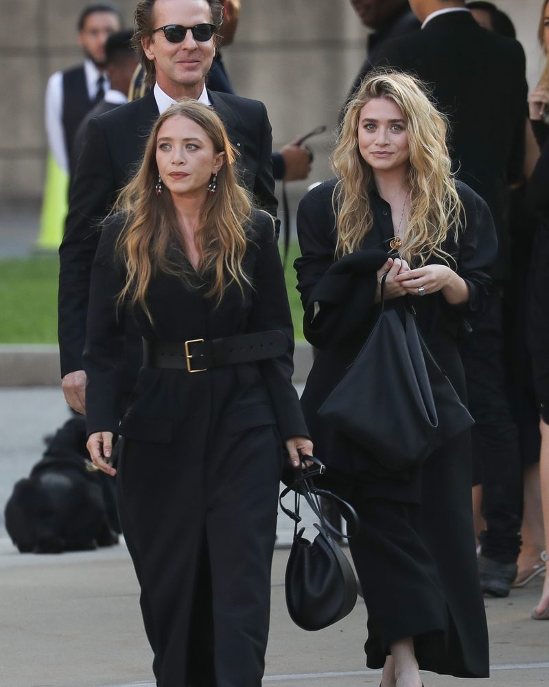 Two women dressed in elegant black outfits walk side by side at an outdoor event%2C each carrying stylish black handbags. They are accompanied by a man in a dark suit and sunglasses in the background. The scene captures a chic%2C fashion-forward moment with coordinated monochrome styling.Two women dressed in elegant black outfits walk side by side at an outdoor event%2C each carrying stylish black handbags. They are accompanied by a man in a dark suit and sunglasses in the background. The scene captures a chic%2C fashion-forward moment with coordinated monochrome styling.