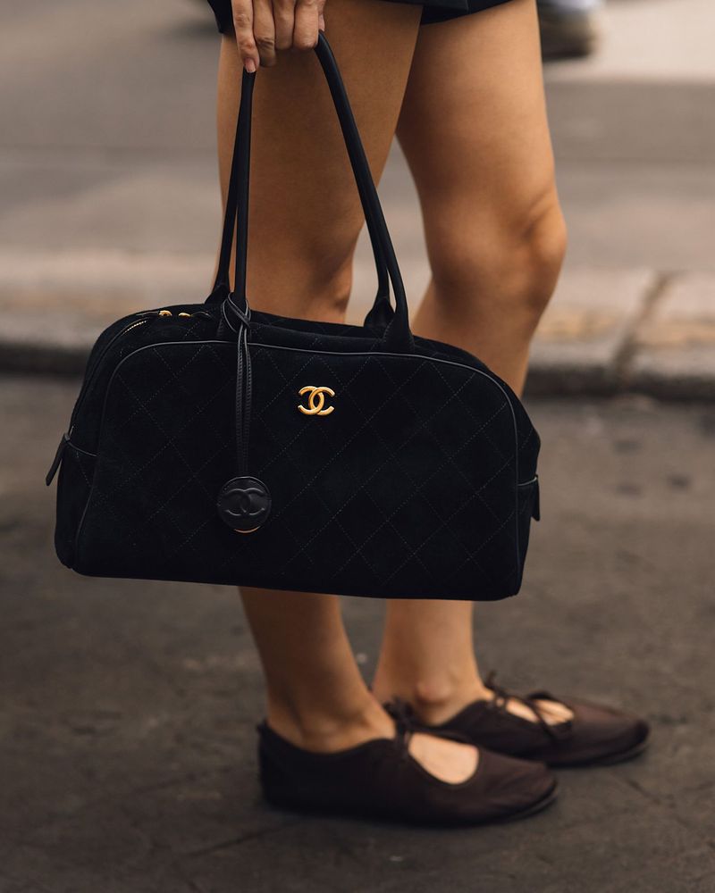 Black quilted Chanel handbag with gold logo hardware held by a person wearing black shorts and brown ballet flats