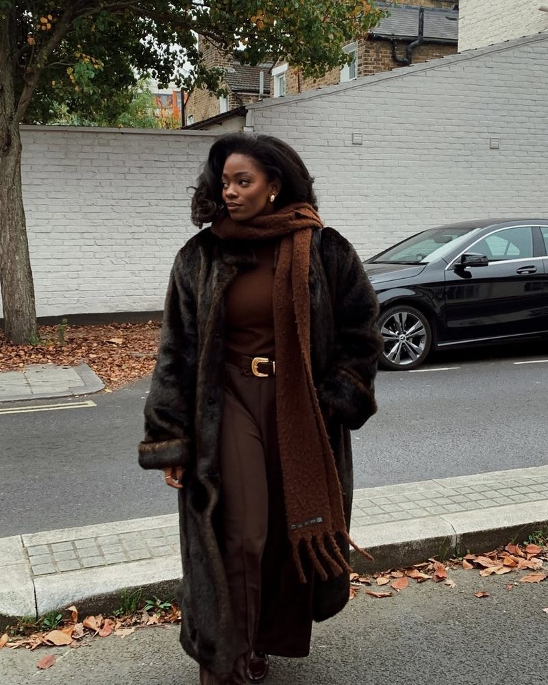 A woman wearing a long dark brown faux fur coat over a turtleneck and wide-leg pants walks along a leaf-strewn city street%2C styled with a matching knit scarf for a chic fall look.