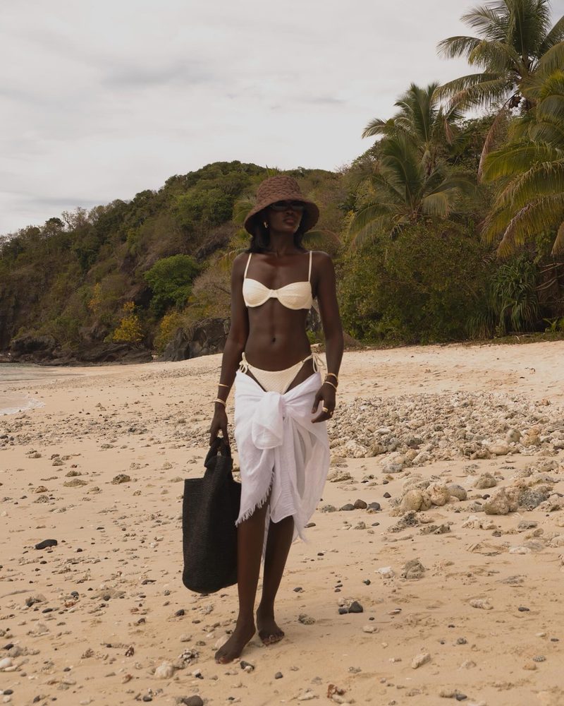 Sun-kissed model in a cream bikini top%2C white sarong%2C and a textured brown bucket hat%2C walking along a rocky beach with palm trees