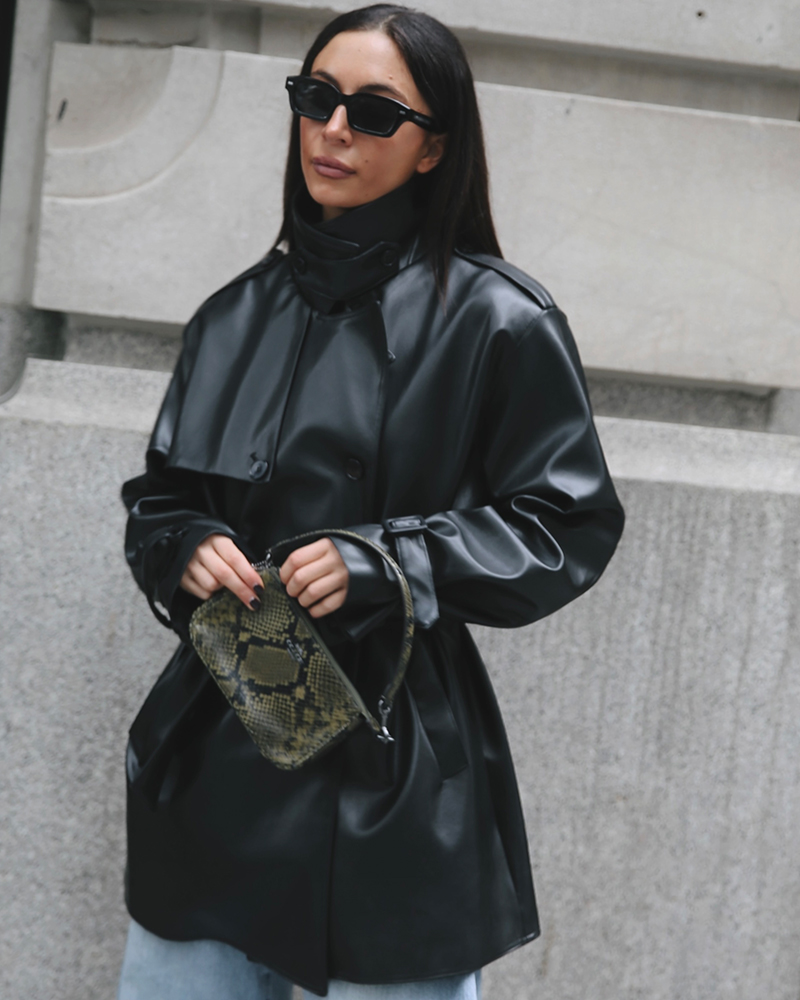 Sleek street-style look featuring a black faux-leather trench%2C dark sunglasses and a snakeskin mini bag against a stone city backdrop