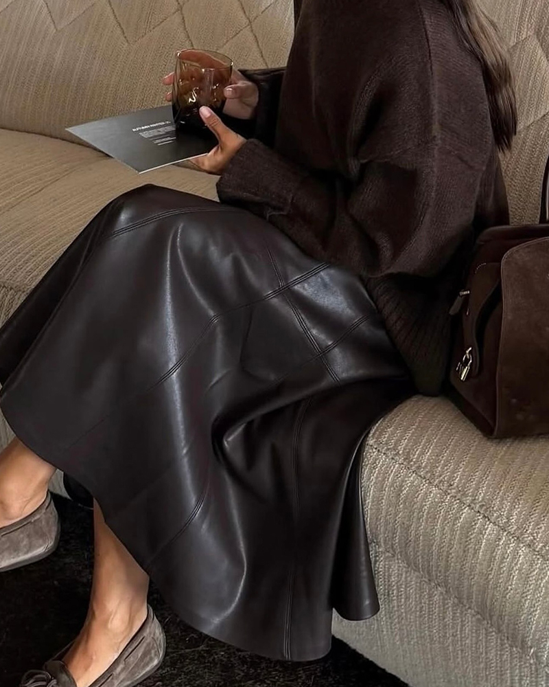 Brown knit sweater paired with a black leather midi skirt and suede loafers%2C styled with a warm drink and handbag on a textured neutral sofa