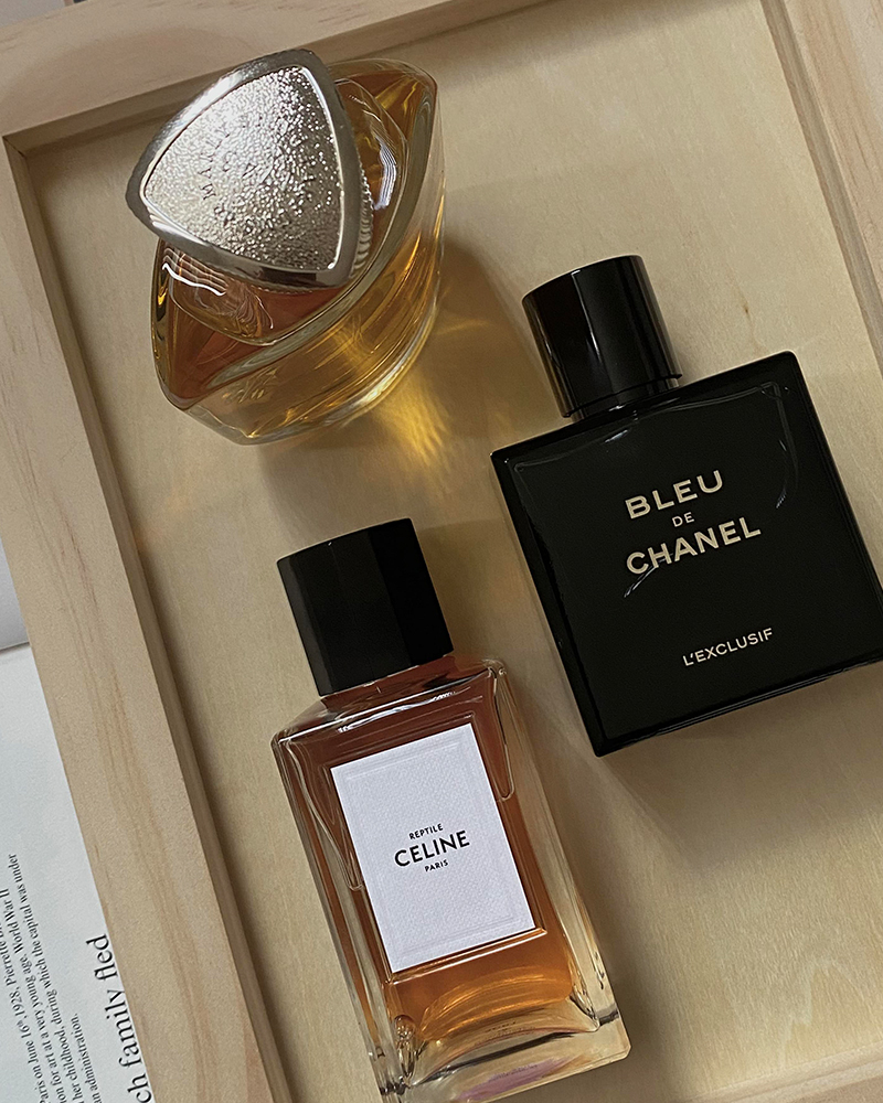 Luxury fragrance trio featuring Celine Reptile and Bleu de Chanel bottles arranged with a sculptural gold-topped perfume on a wooden tray