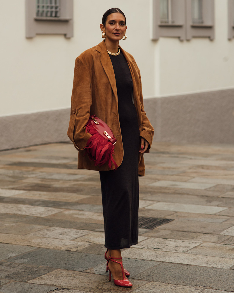 Street style look featuring a tan suede blazer over a black maxi dress%2C paired with red feathered bag%2C red heels%2C and gold statement jewelry
