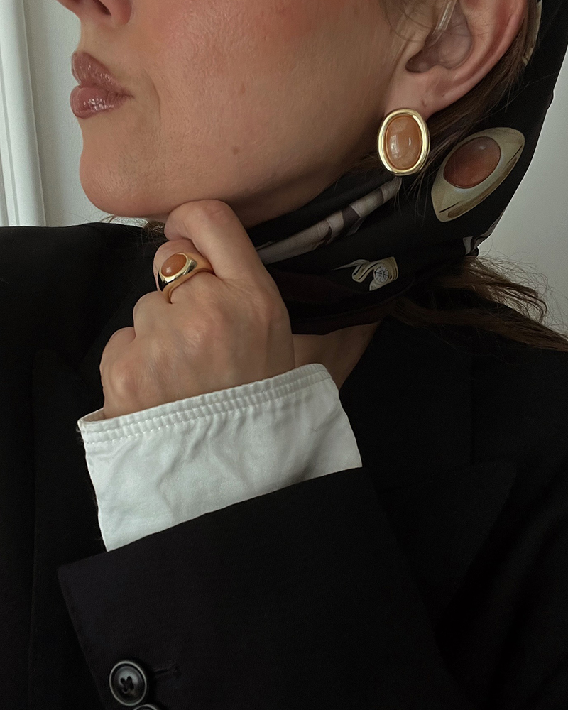 Close-up of polished gold statement earrings and matching gold ring styled with a black blazer%2C white cuff%2C and patterned silk scarf