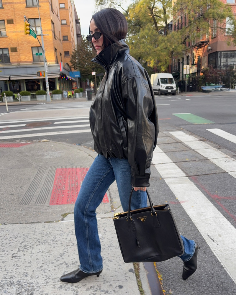 Sleek street style featuring a black leather funnel-neck jacket%2C straight-leg jeans%2C and a black Prada top-handle bag while crossing a city street