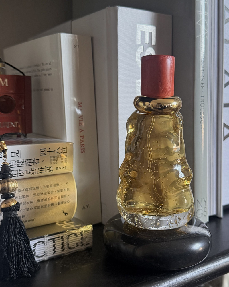 Bottega Veneta perfume bottle with amber glass and leather cap styled on a bookshelf among art and fashion books