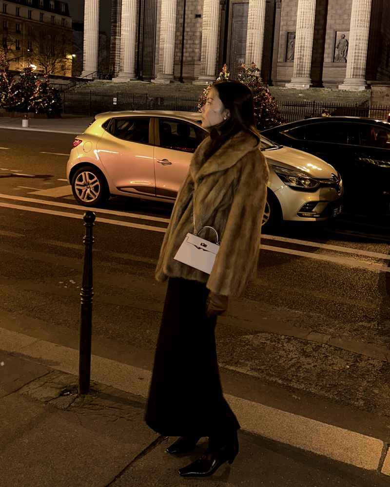 Fashion-forward figure in a faux-fur coat carrying a small white top-handle bag on a city street at night near festive lights