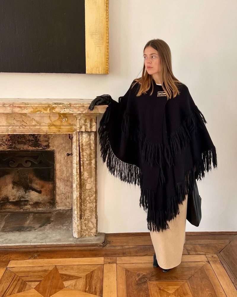 Chic winter outfit featuring a black fringe poncho draped over a light beige maxi skirt%2C complemented by stylish black gloves and a sleek handbag.