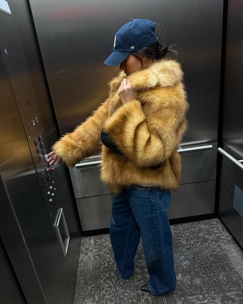 Casual chic look featuring a model in a luxurious faux fur coat paired with wide-leg jeans and a navy baseball cap%2C set in an elevator.