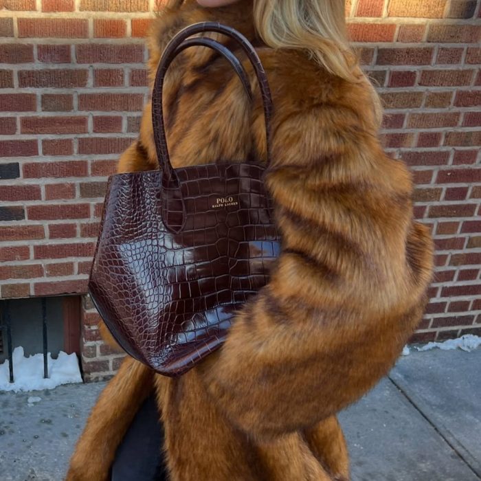 A luxurious brown fur coat paired with a stylish burgundy crocodile-embossed tote from Polo Ralph Lauren, set against a brick backdrop.