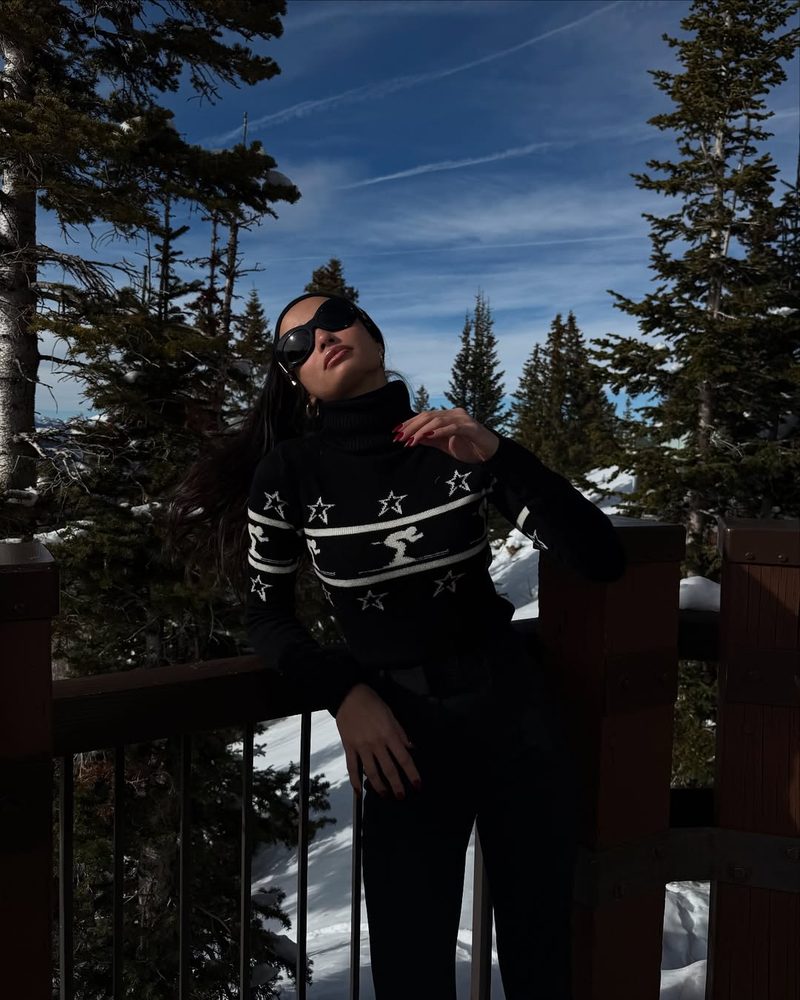 Moncler sweater features a black base adorned with white skiing motifs and stars%2C paired with sleek black pants and oversized sunglasses