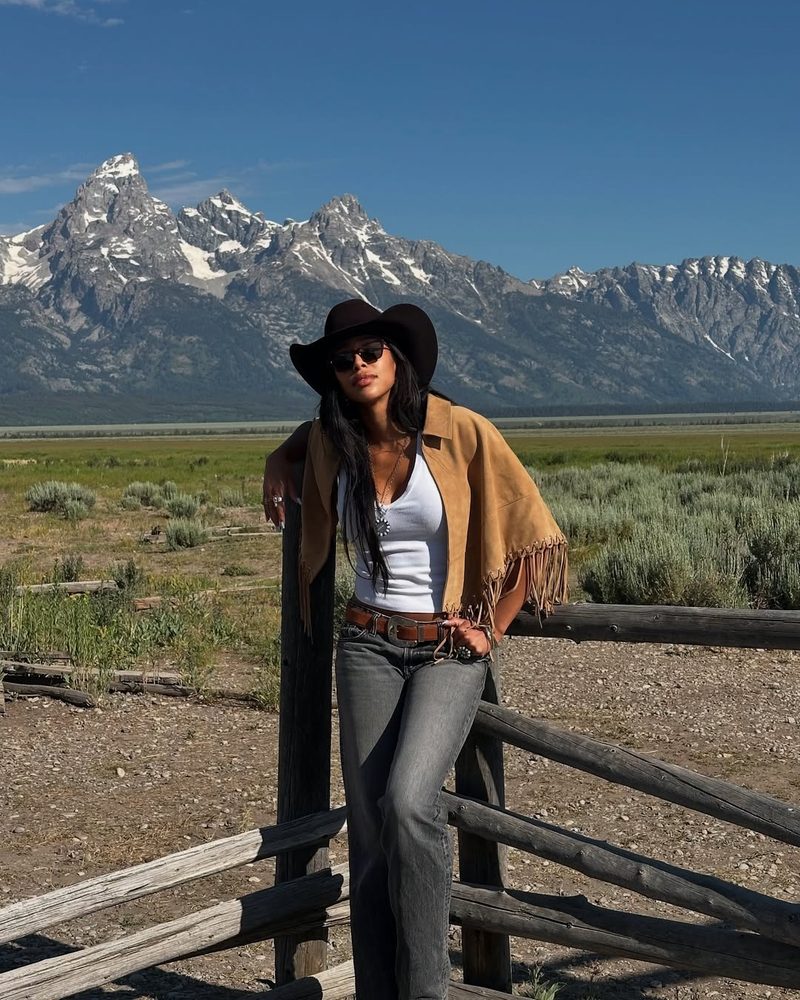 Western-inspired outfit features a tan fringed poncho paired with a white tank top and flared gray jeans%2C accentuated by a wide-brimmed hat and sunglasses