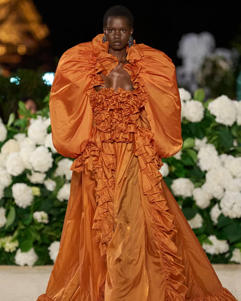 Model walking the runway in a dramatic burnt orange ruffled evening gown with voluminous sleeves%2C set against a floral nighttime backdrop