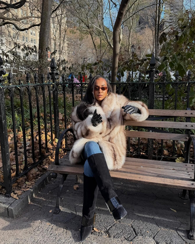Woman in plush beige fur coat with black leather gloves%2C knee-high boots%2C and sunglasses%2C seated on a park bench in winter greenery