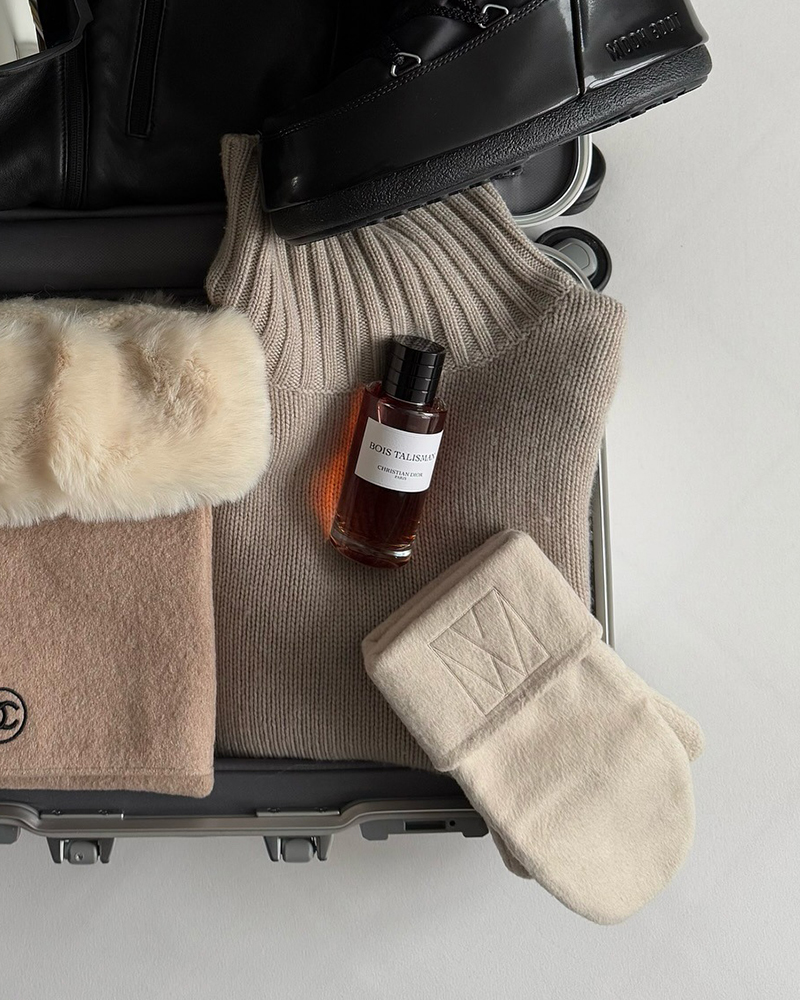 Cashmere mittens styled with knit sweater%2C Dior Bois Talisman perfume and leather boots in a packed travel suitcase