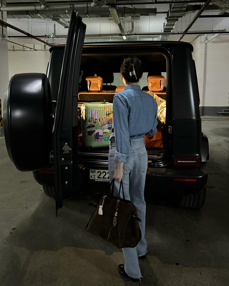 Woman loading colorful luggage into a black SUV%2C wearing a denim shirt and jeans with a brown leather tote%2C inside a parking garage