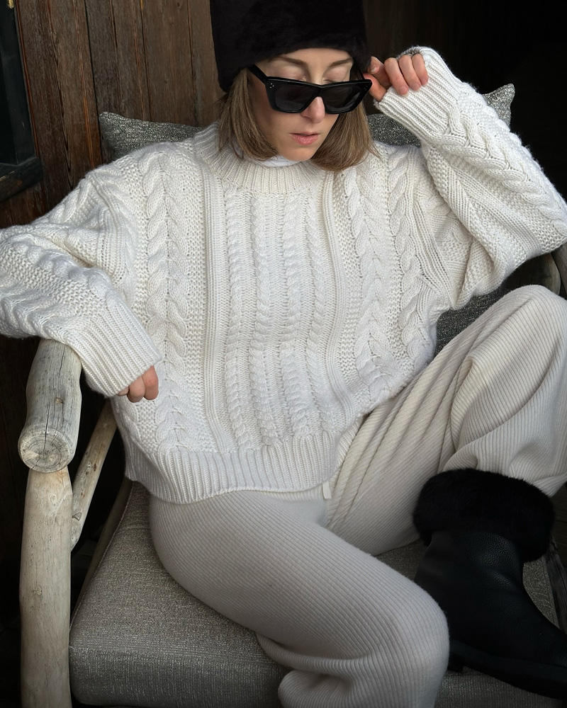 Cozy winter look featuring a cream cable-knit sweater and ribbed lounge pants with beanie and sunglasses on a rustic chair