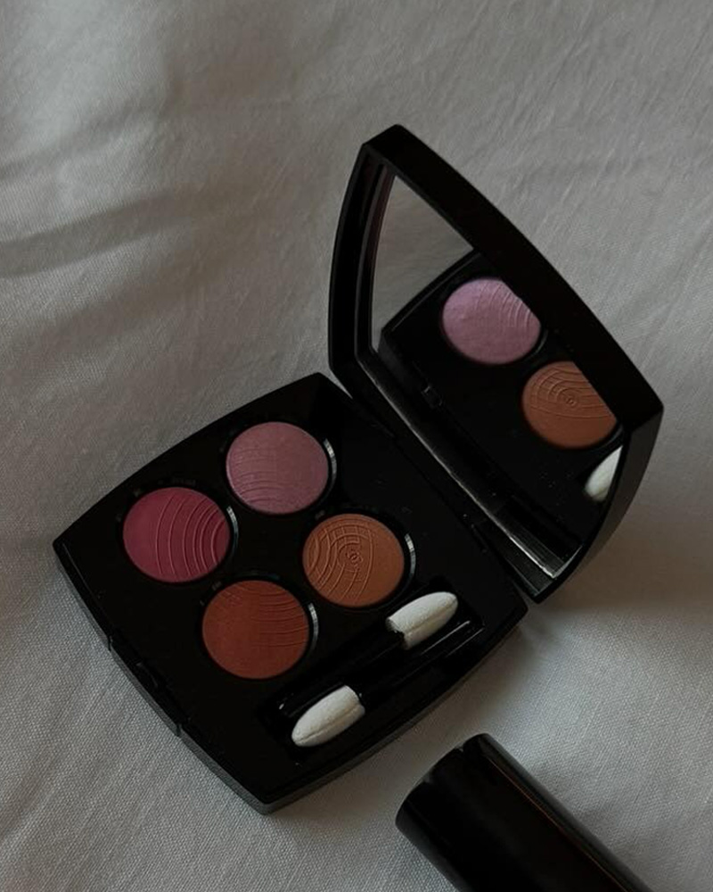 Open eyeshadow palette with mauve and bronze shades%2C dual applicators and black compact%2C styled on soft neutral fabric backdrop