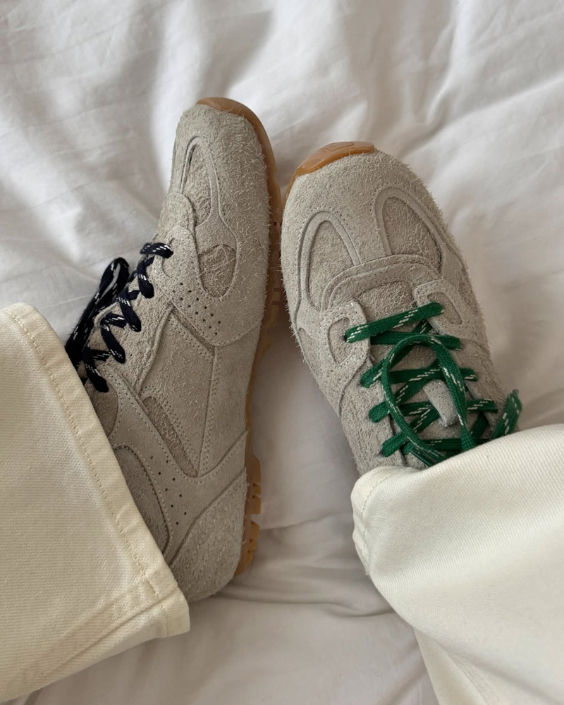 Close-up of gray suede sneakers with contrasting black and green laces%2C styled with white pants on a soft white bed backdrop
