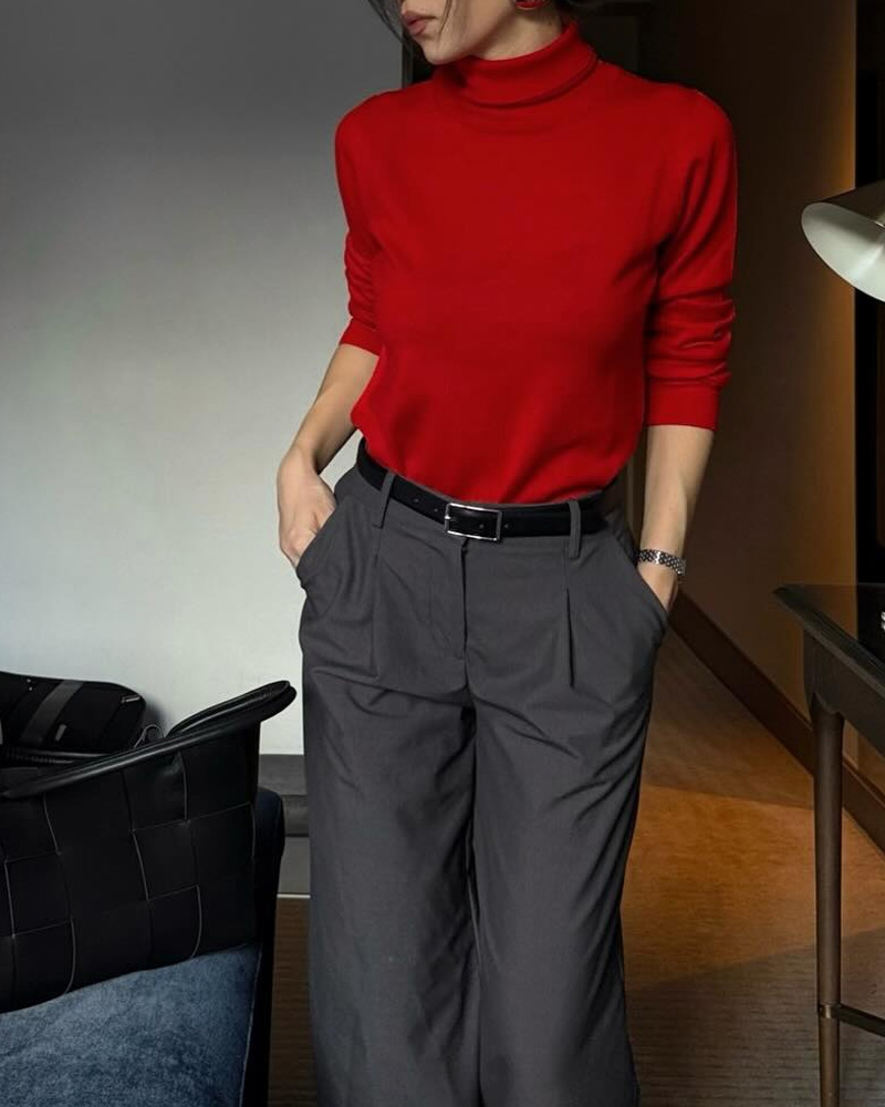 Model styled in a red turtleneck sweater with gray tailored trousers and a slim black leather belt%2C indoors