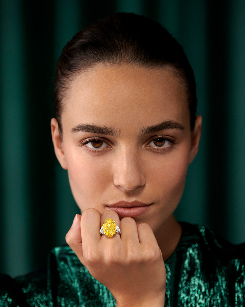 Close-up of a model showcasing a large yellow diamond ring with white diamond sides%2C styled against deep green drapery for a luxe holiday look