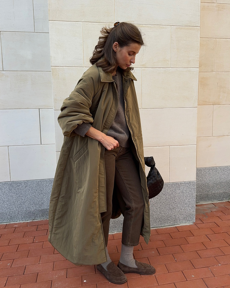 Neutral fall outfit featuring an oversized olive trench%2C taupe knit sweater%2C moccasins%2C and a woven black bag%2C styled against a stone backdrop