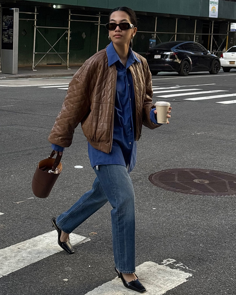 Street style look with quilted brown jacket%2C blue shirt%2C straight-leg jeans%2C leather bucket bag%2C crossing a city street