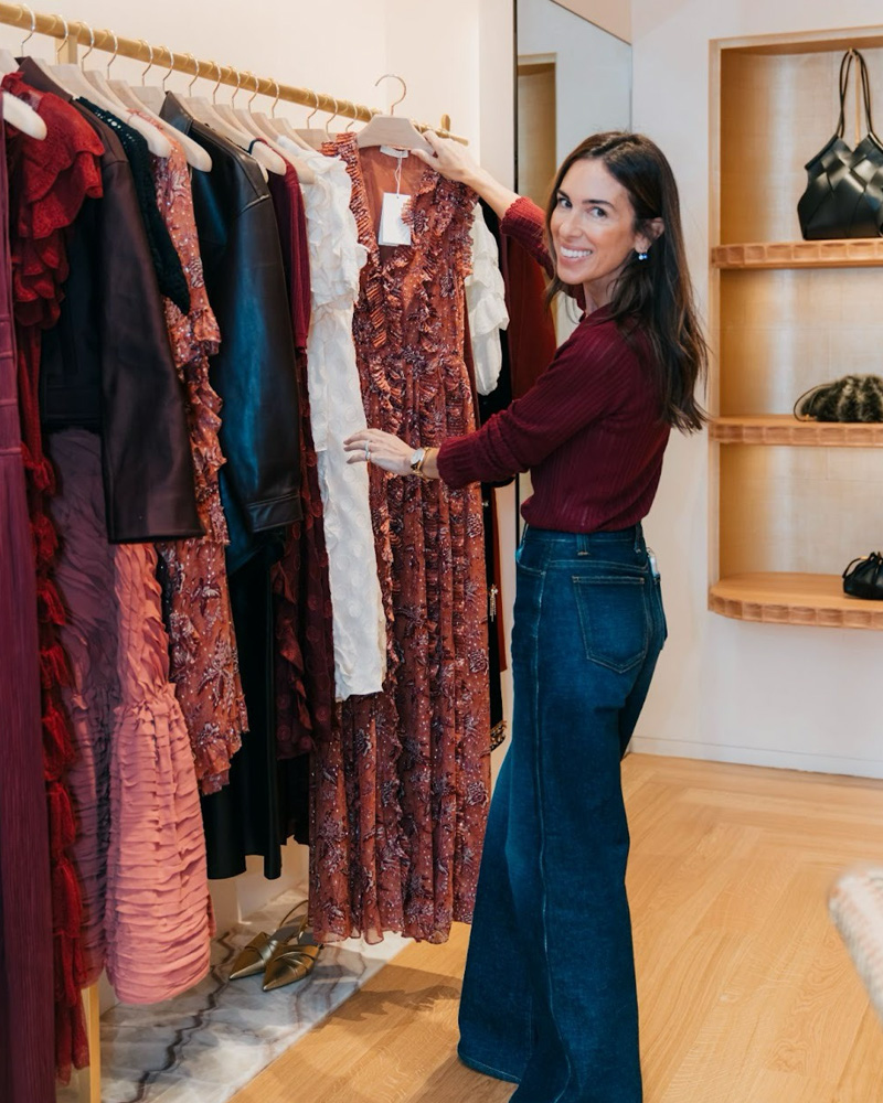 Stylist browsing embellished Ulla Johnson dresses while wearing a burgundy sweater and wide-leg denim inside a modern boutique showroom