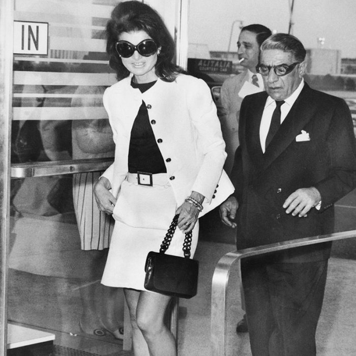 A woman in a stylish white suit and large sunglasses walks confidently through a glass door, followed by an older man in a dark suit. They exude elegance.