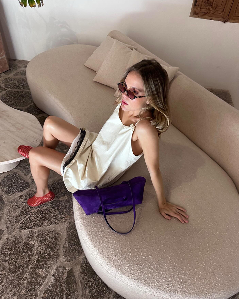 Purple suede tote bag paired with a cream camisole mini dress%2C tortoiseshell sunglasses and red flats in a minimalist living room setting