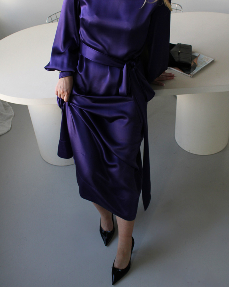 Model wearing a deep purple satin midi dress with belted waist and black pointed heels%2C styled in a minimalist studio interior