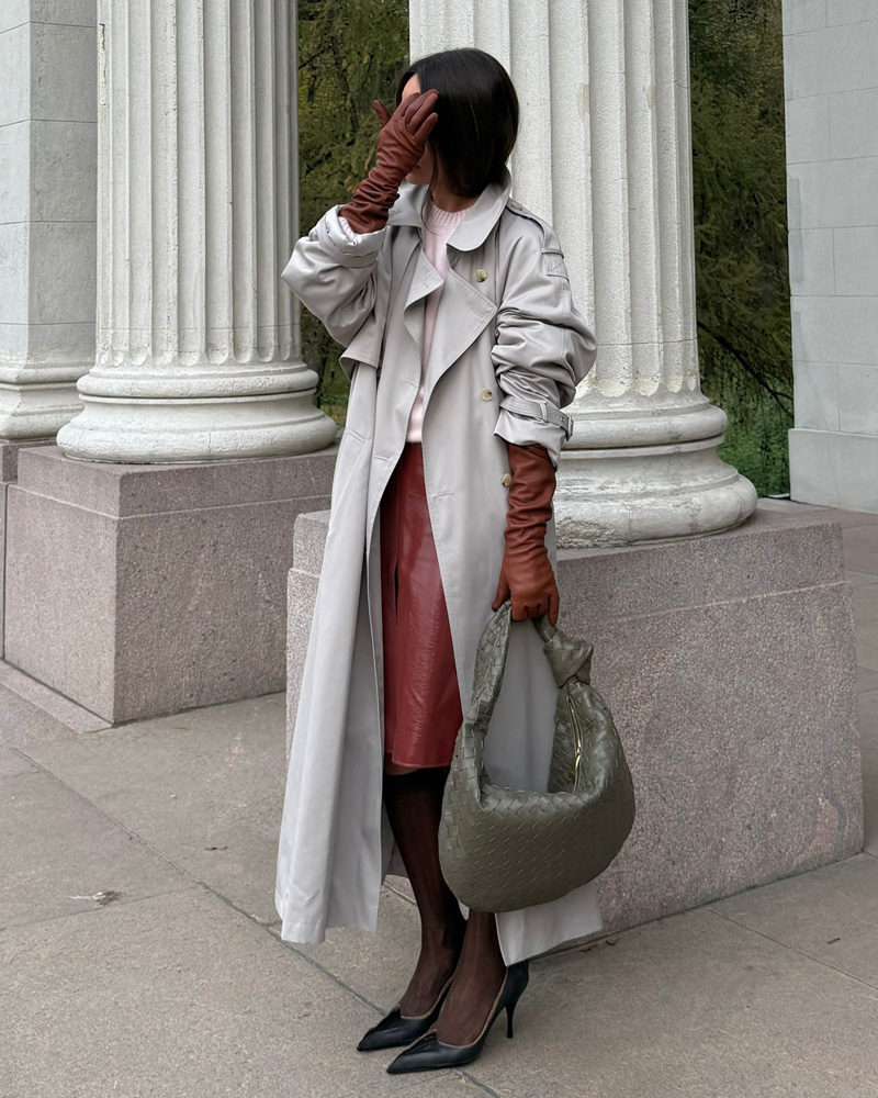 Street style look wearing a light trench coat%2C burgundy leather skirt%2C opera gloves and woven hobo bag by stone columns