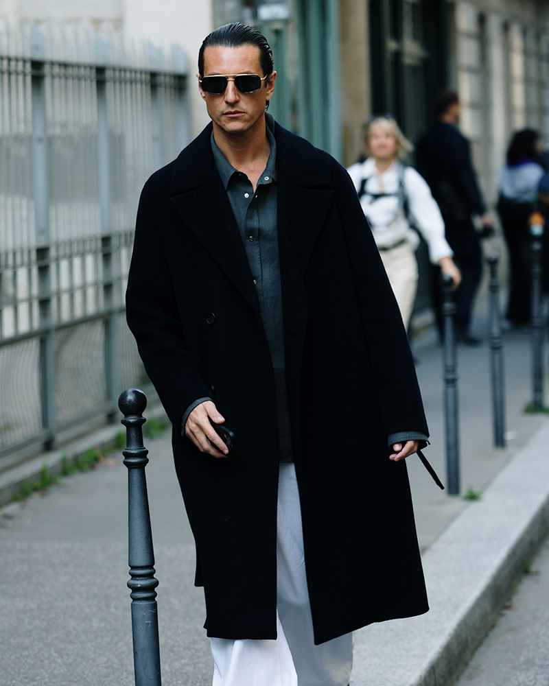 Sharp city look featuring a man in a tailored black overcoat%2C charcoal button-down shirt and sleek aviator sunglasses walking through an urban street backdrop