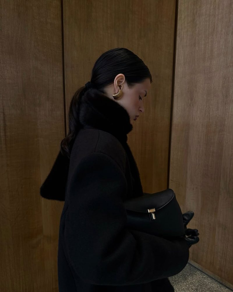 Chic winter street style with a black wool coat%2C gold hoop earrings and structured leather bag against wood panel walls