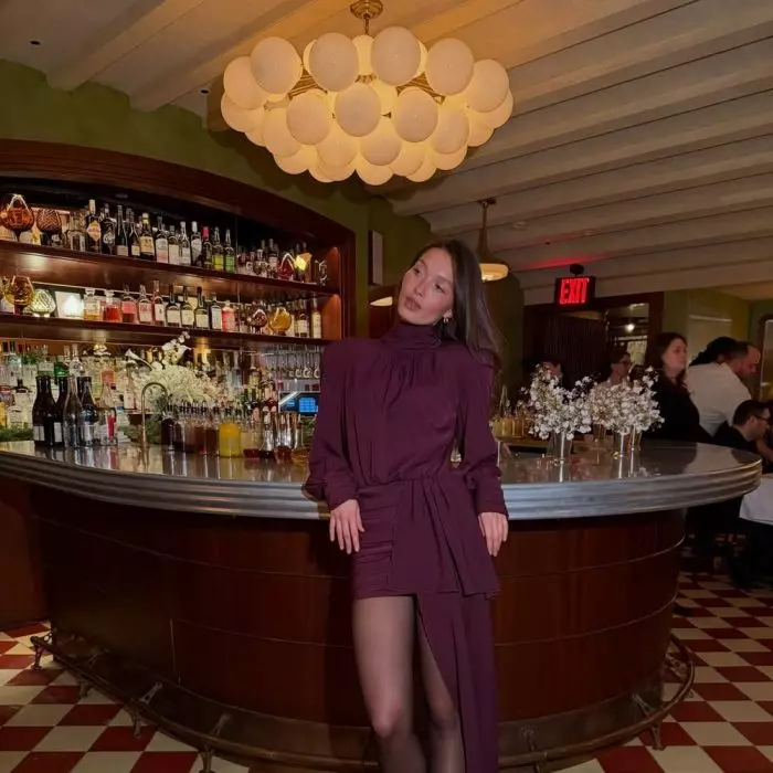 Chic burgundy wrap dress with gathered detailing and an asymmetrical hem, paired with sheer black tights and classic black heels in a vibrant bar setting.