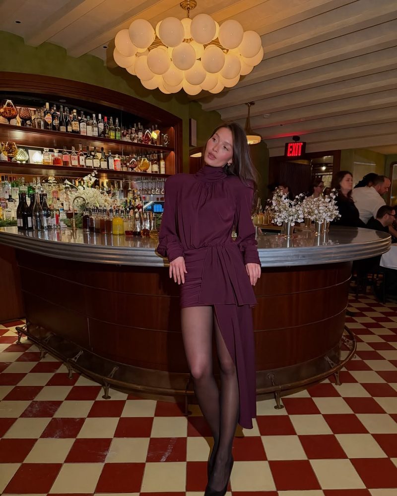 Chic burgundy wrap dress with gathered detailing and an asymmetrical hem%2C paired with sheer black tights and classic black heels in a vibrant bar setting