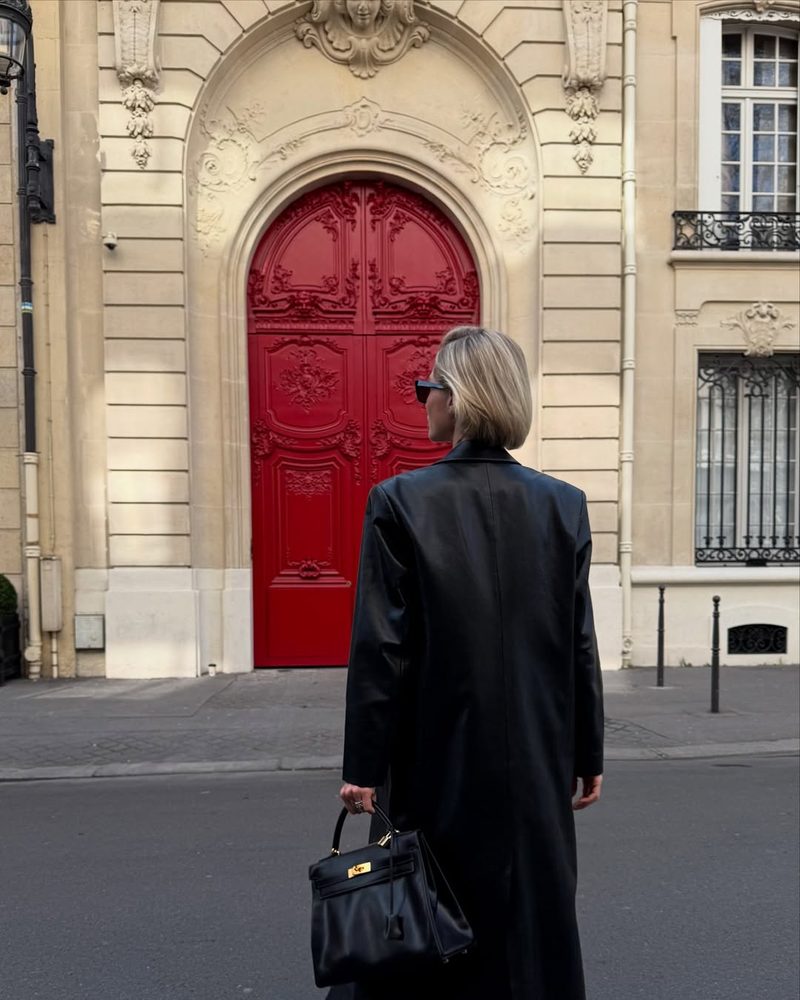 Chic black leather coat paired with oversized sunglasses%2C elegantly holding a structured black handbag in front of a vibrant red door