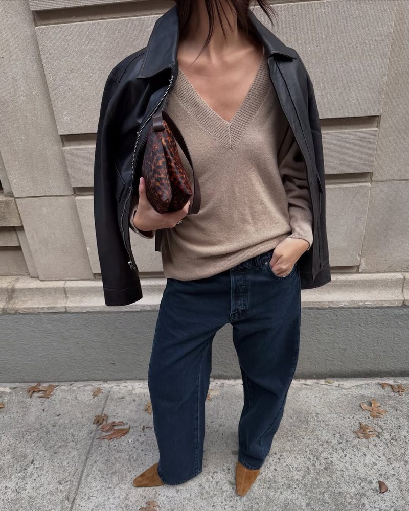 Black leather jacket layered over a beige V-neck sweater with wide-leg jeans%2C suede heels%2C and a brown patterned clutch for chic street style